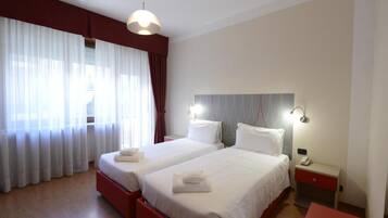 Quarto de hotel com duas camas, uma parede de destaque vermelha, uma janela grande com cortinas leves e uma mesa de cabeceira com abajur.