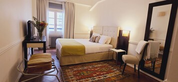 Quarto de hotel com uma cama, uma escrivaninha, uma cadeira, um espelho e uma janela com cortinas.