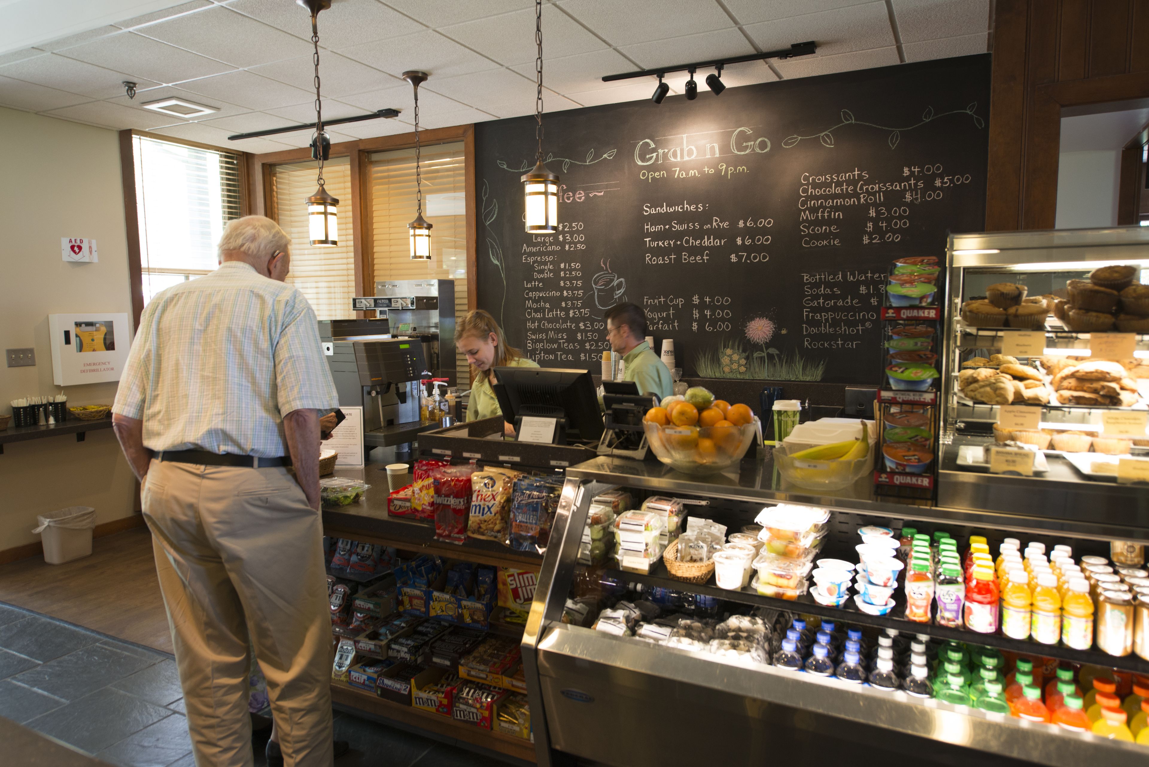 Interior de cafeteria com quadro de cardápio, caixa registradora e diversos alimentos expostos.