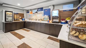 Uma área de café da manhã moderna com estação de autoatendimento, refrigerador para iogurtes e uma cesta de frutas.