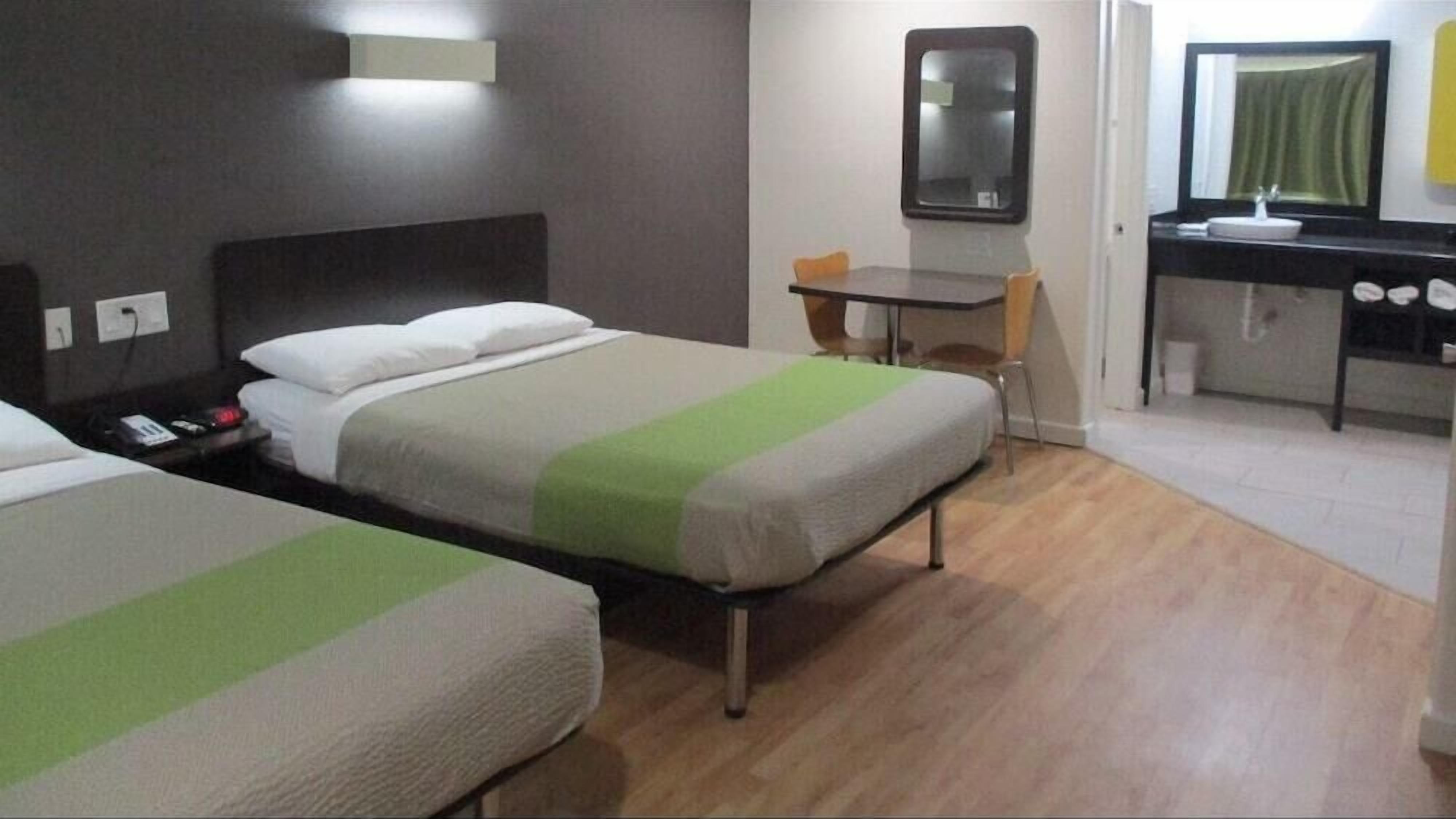Quarto de hotel com duas camas, uma escrivaninha, uma cadeira, um espelho e banheiro com pia e vaso sanitário.