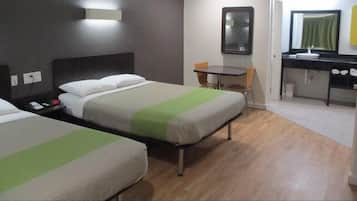 Quarto de hotel com duas camas, uma escrivaninha, uma cadeira, um espelho e banheiro com pia e vaso sanitário.