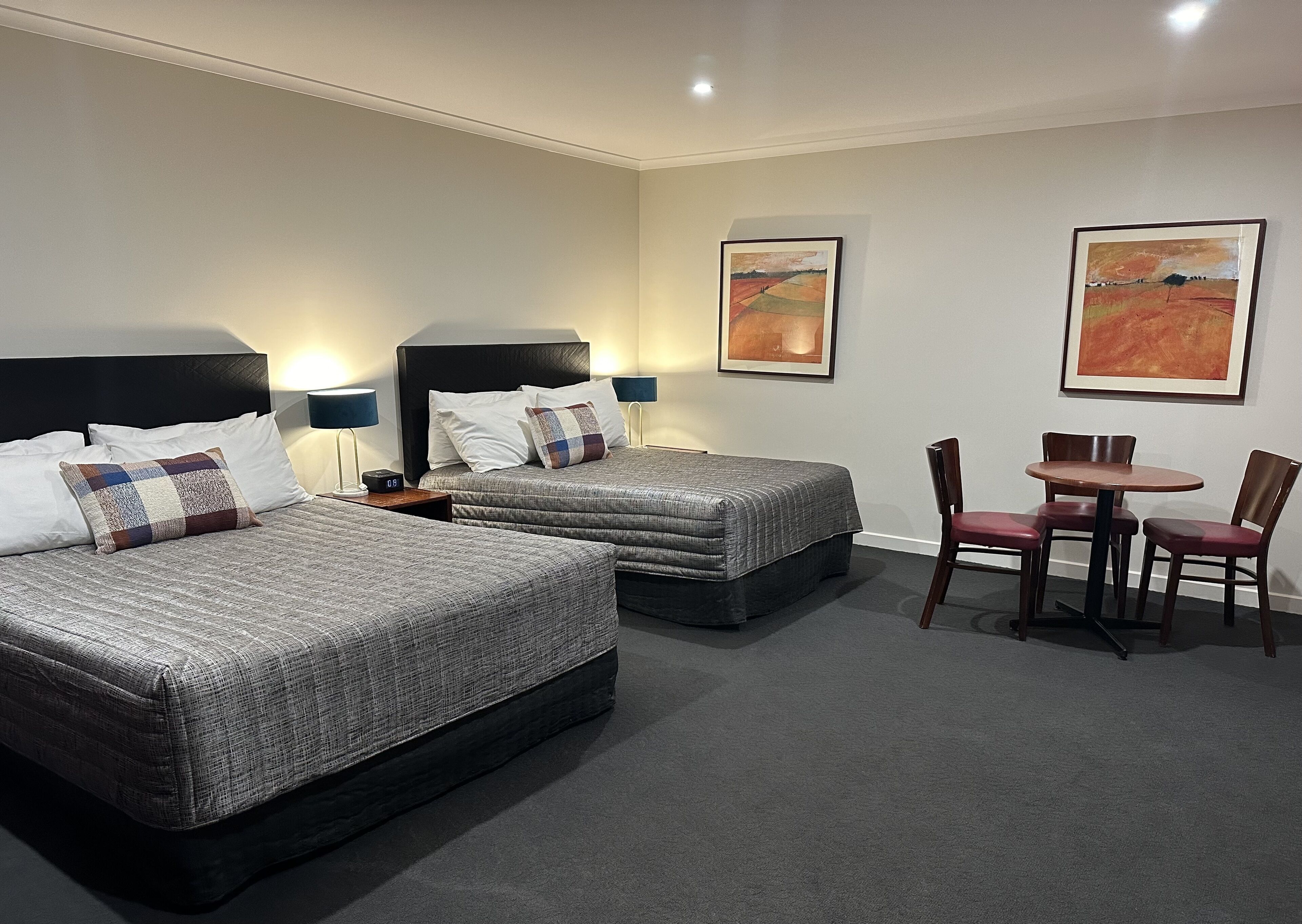 Quarto de hotel com duas camas, uma mesinha com cadeiras e dois quadros na parede.