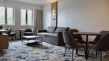 Quarto de hotel moderno com sofá, duas poltronas, uma mesinha e uma janela grande.