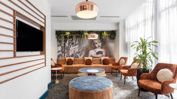Uma área de estar moderna com um amplo sofá, pufes redondos e poltronas. Uma televisão de tela plana está fixada na parede, e um mural é visível ao fundo.