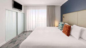 Quarto de hotel com uma cama grande, televisão de tela plana na parede, uma janela ampla com cortinas translúcidas e cabeceira com painel estofado em tom claro.