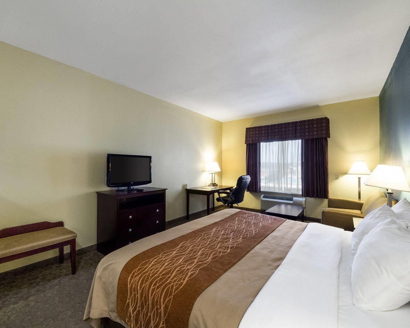Quarto de hotel com cama, escrivaninha, cadeira, televisão e janela com cortinas.
