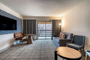 Quarto de hotel com uma televisão de tela grande, um sofá, uma poltrona, uma mesinha e um tapete estampado.