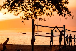 Jogo de voleibol de praia ao pôr do sol, com rede e jogadores em silhueta contra o céu.
