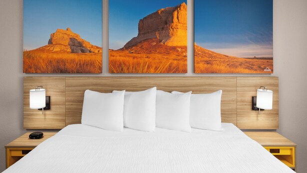 Quarto de hotel com uma cama grande, roupa de cama branca, duas mesas de cabeceira com abajures e uma obra de arte na parede retratando uma paisagem desértica.