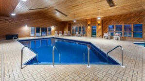 Piscina interior com teto de madeira e chão de azulejos, rodeada por espreguiçadeiras e janelas.