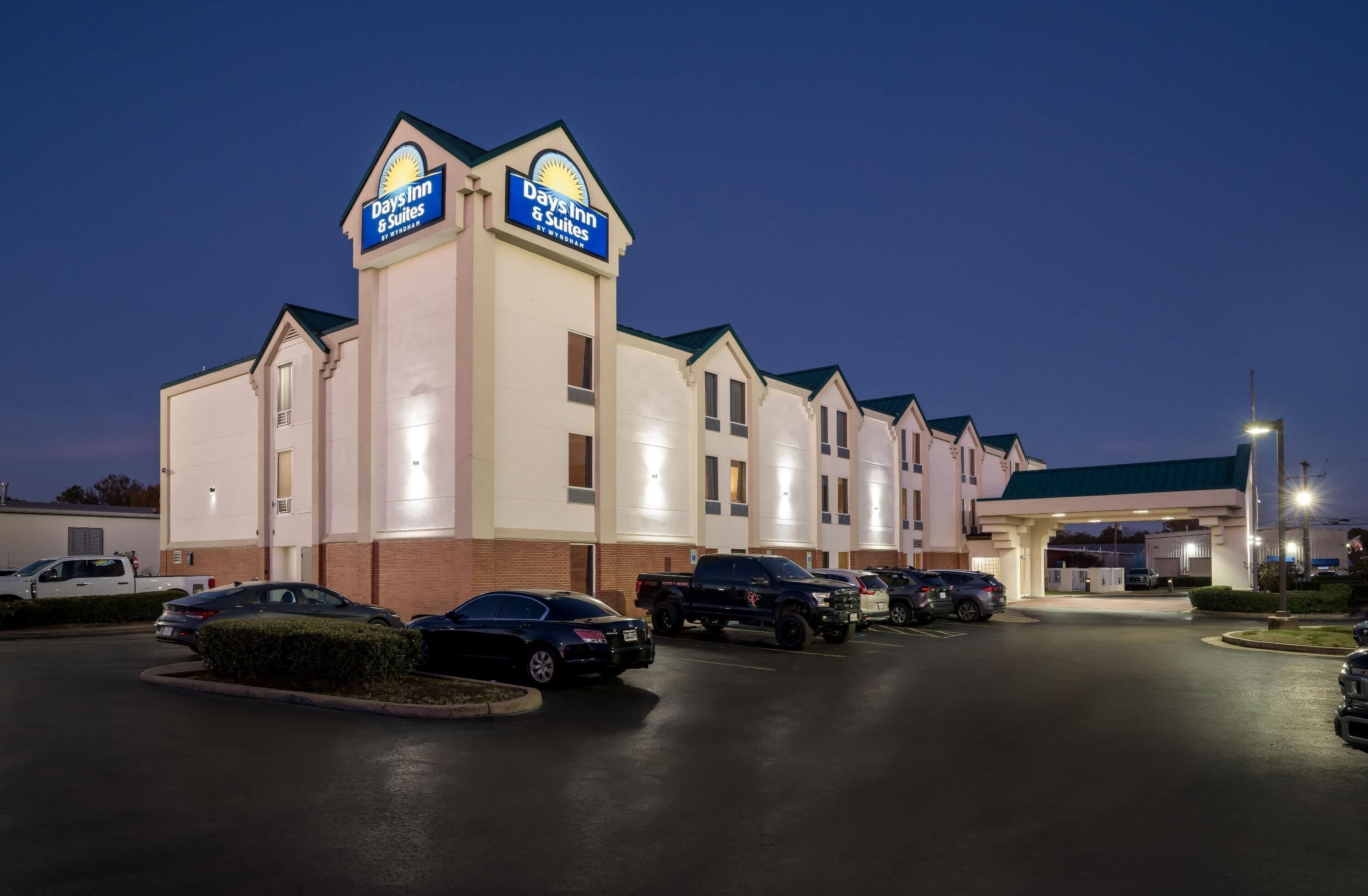 Um hotel Days Inn & Suites com vários edifícios, um estacionamento com diversos carros e uma entrada bem iluminada.