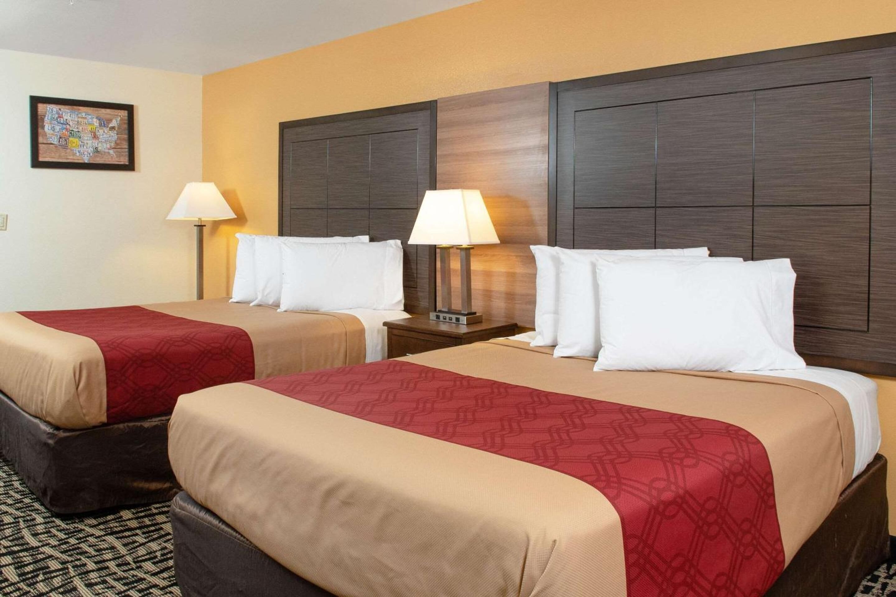 Quarto de hotel com duas camas, cada uma com colcha vermelha e bege, duas luminárias de cabeceira e uma mesinha de cabeceira entre as camas.