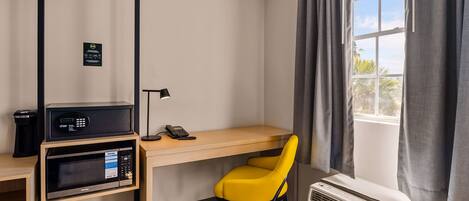 Quarto de hotel compacto com escrivaninha, micro-ondas, cadeira, telefone e ar condicionado.