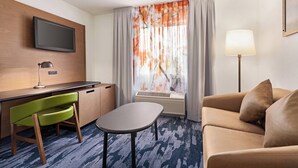 Quarto de hotel com uma escrivaninha, uma cadeira verde, um sofá, uma televisão fixada na parede, uma janela com cortinas e um abajur.