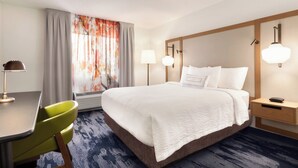 Quarto de hotel com cama, mesa de cabeceira, abajur, cadeira e janela com cortinas.