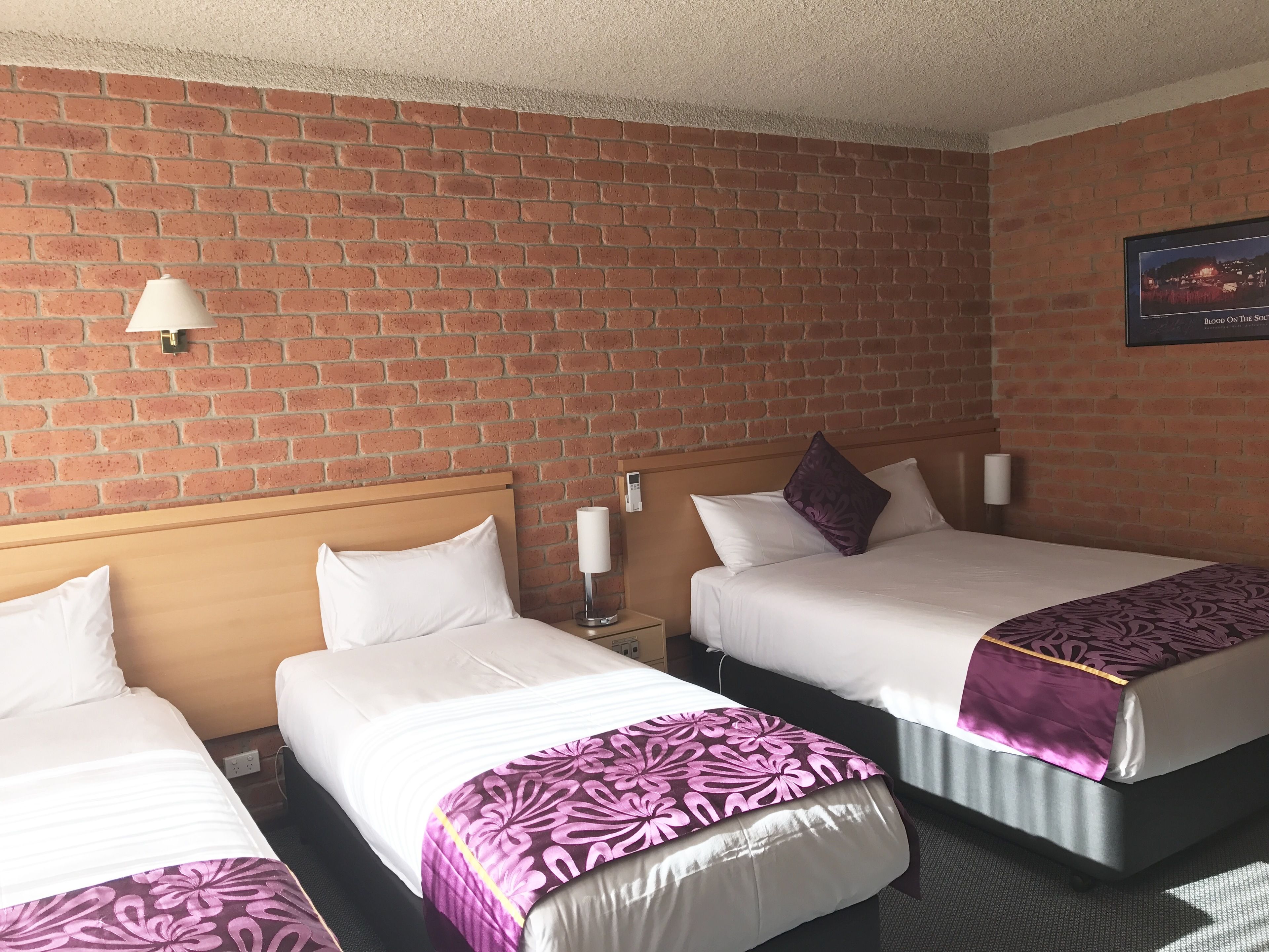 Quarto de hotel com duas camas, parede de tijolos, quadro emoldurado e mesa de cabeceira com abajur.