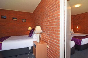 Quarto de hotel com paredes de tijolo, duas camas, uma mesinha de cabeceira com abajur e uma pequena mesa.