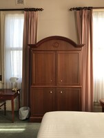 Quarto com um guarda-roupa de madeira grande, uma cama, uma escrivaninha com abajur e cortinas leves.