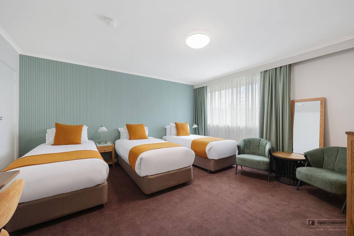 Quarto de hotel com três camas, uma poltrona verde, uma mesinha e um espelho grande.