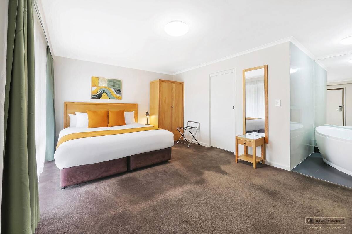 Quarto de hotel com uma cama grande, um guarda-roupa de madeira, uma mesa pequena, um espelho e uma casa de banho com banheira e sanita.