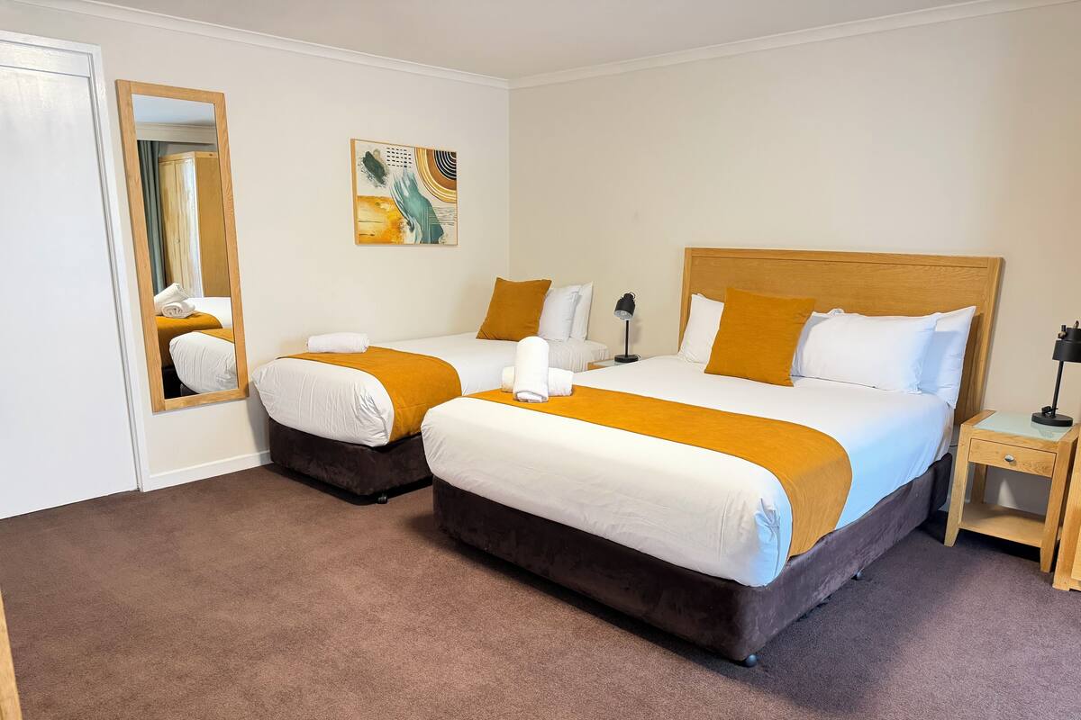 Quarto de hotel com duas camas, cabeceira de madeira, espelho, mesa de cabeceira e um quadro na parede.