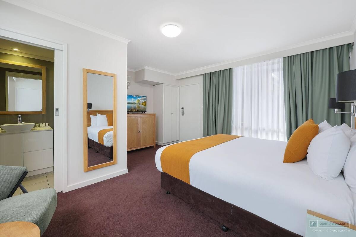 Quarto de hotel com uma cama grande, uma escrivaninha, uma cadeira, um espelho, uma televisão e um guarda-roupa.