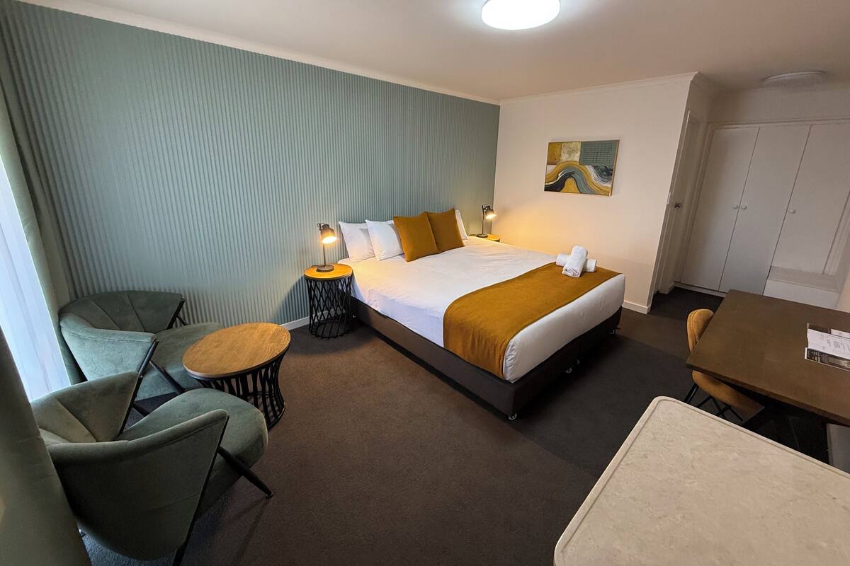 Quarto de hotel com uma cama, duas cadeiras, uma mesa redonda pequena, uma escrivaninha e um guarda-roupa.