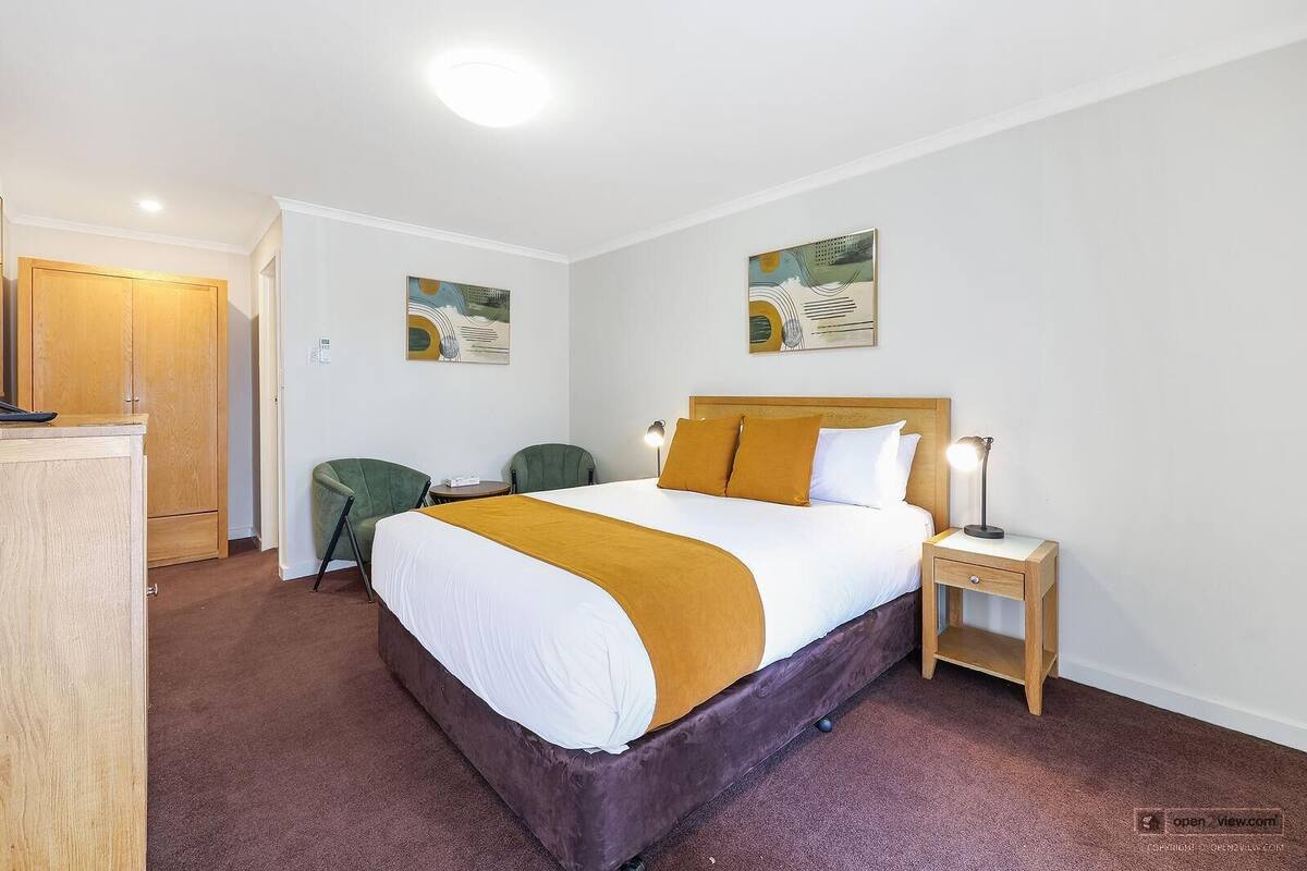 Quarto de hotel com uma cama grande, uma escrivaninha, duas cadeiras, uma mesinha e duas pinturas na parede.