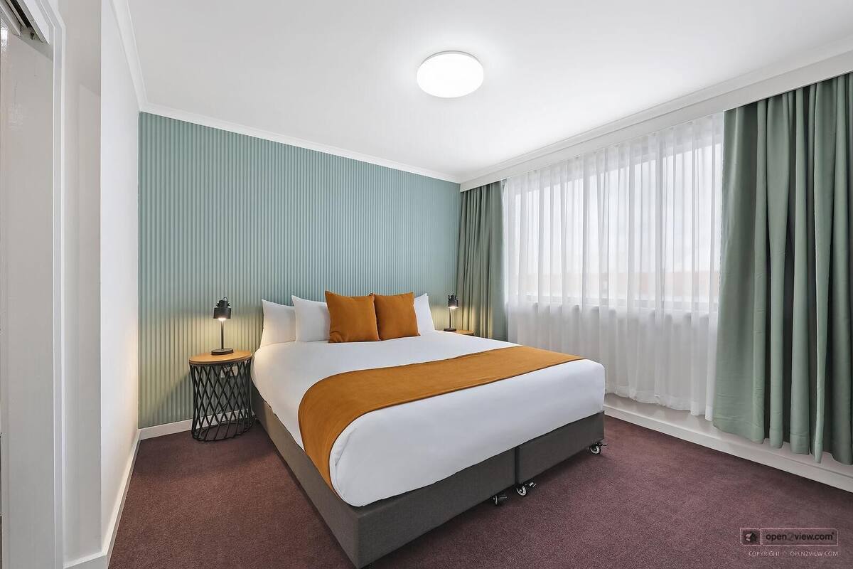 Quarto de hotel com uma cama grande, mesa de cabeceira com abajur e janela com cortinas finas.