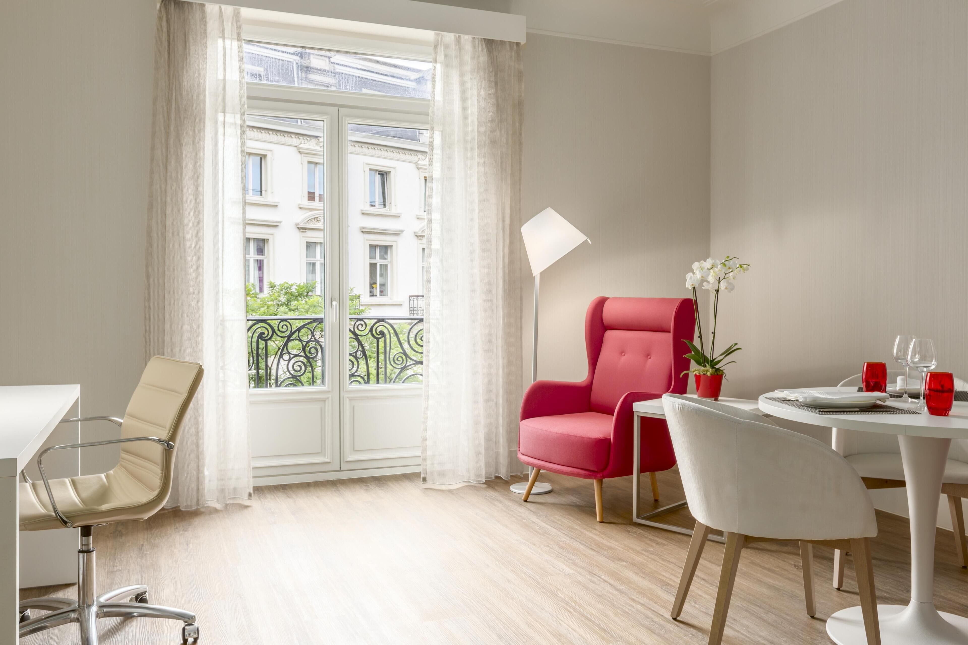 Quarto de hotel moderno com área de refeições, uma poltrona vermelha, uma cadeira branca, uma orquídea em vaso e vista para prédios pela janela.