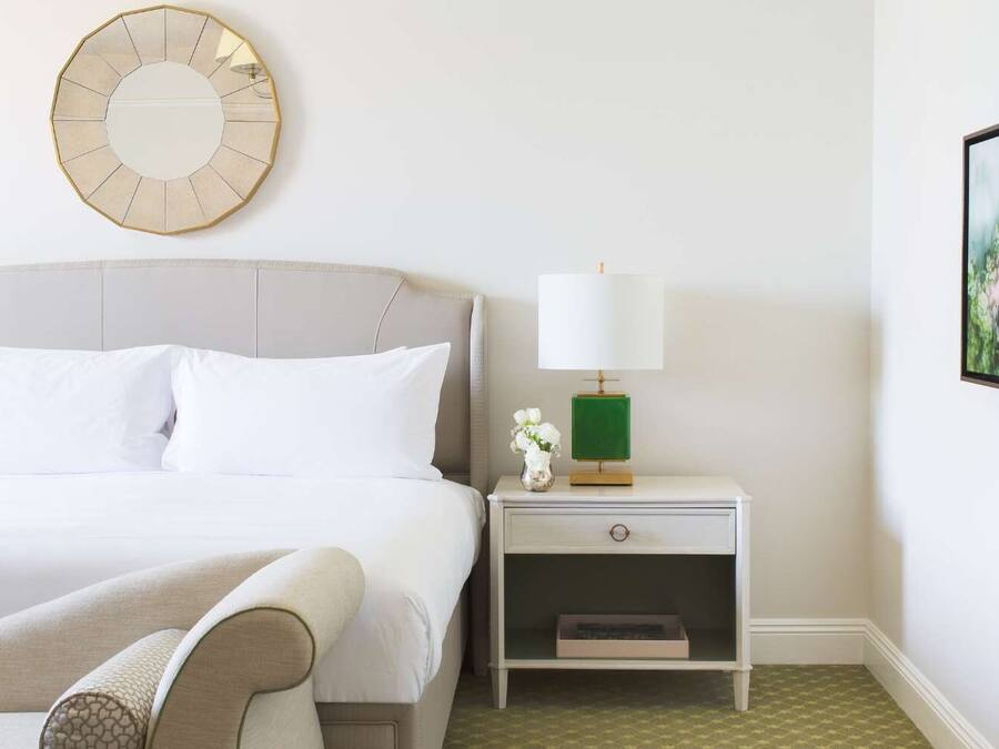 Uma cama bem arrumada com lençóis brancos, uma mesa de cabeceira com abajur verde, um pequeno vaso com flores e um quadro na parede.