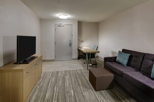 Uma sala de estar moderna com uma TV de tela plana sobre um armário de madeira, um sofá de cor escura com um travesseiro, uma mesa pequena com um abajur e uma porta que dá para outro cômodo.