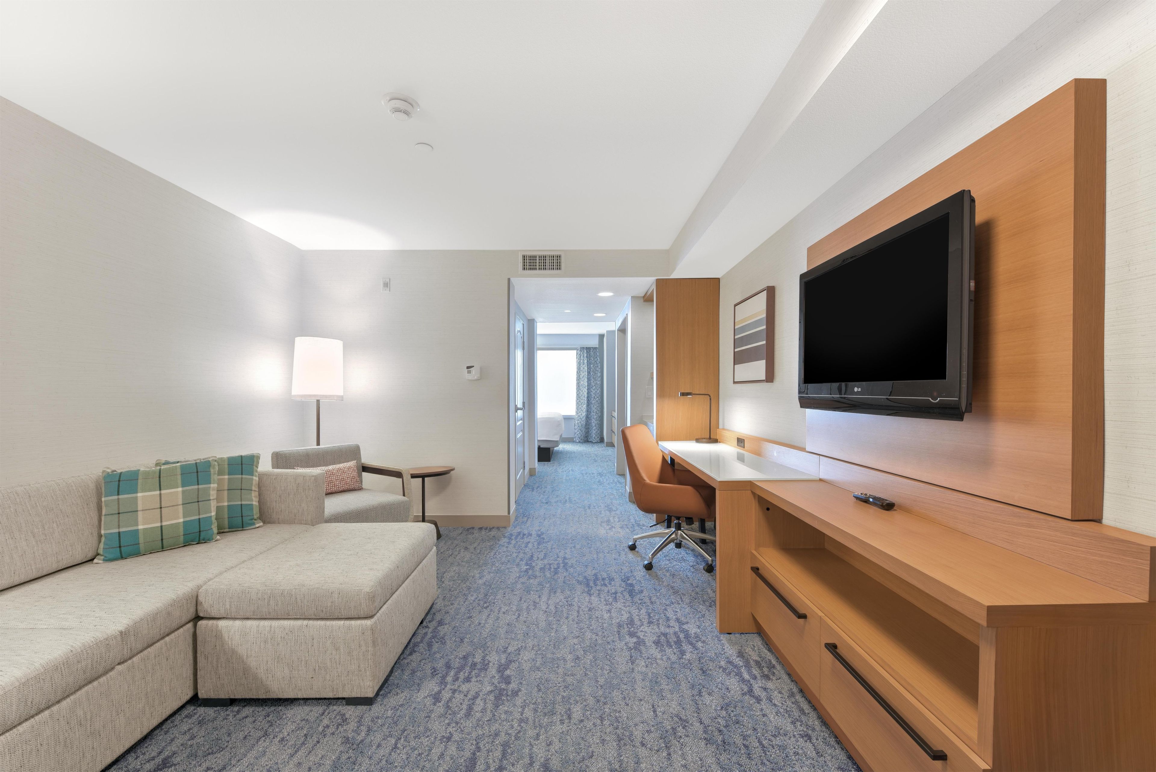 Quarto de hotel com sofá seccional, escrivaninha com televisão de tela plana e pequena mesa com abajur.