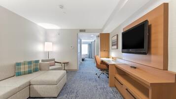 Quarto de hotel com sofá seccional, escrivaninha com televisão de tela plana e pequena mesa com abajur.