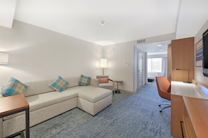Quarto de hotel com sofá seccional, uma pequena mesa, escrivaninha, cadeira e cama ao fundo.