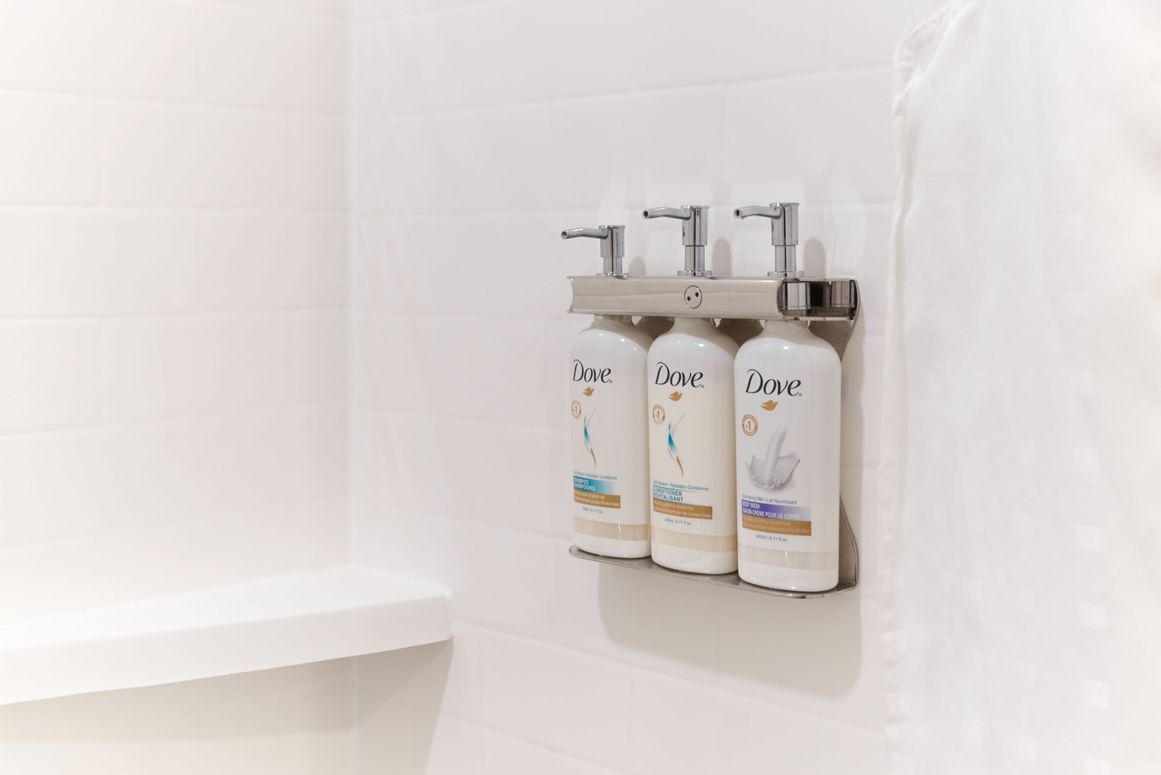 Casa de banho com três dosadores de loção Dove fixados numa parede revestida a azulejos brancos.