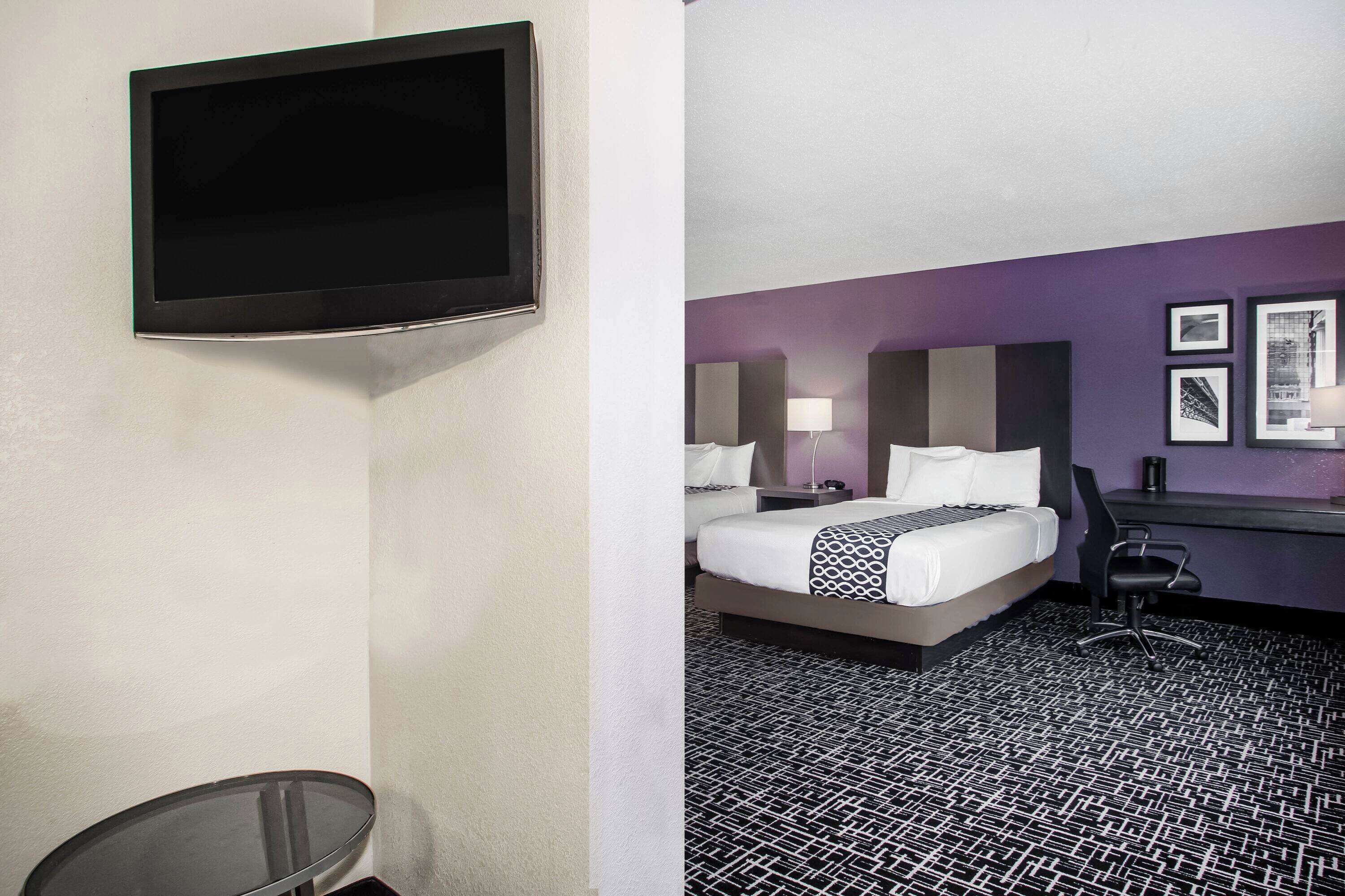 Quarto de hotel com cama, escrivaninha, cadeira e televisão na parede.