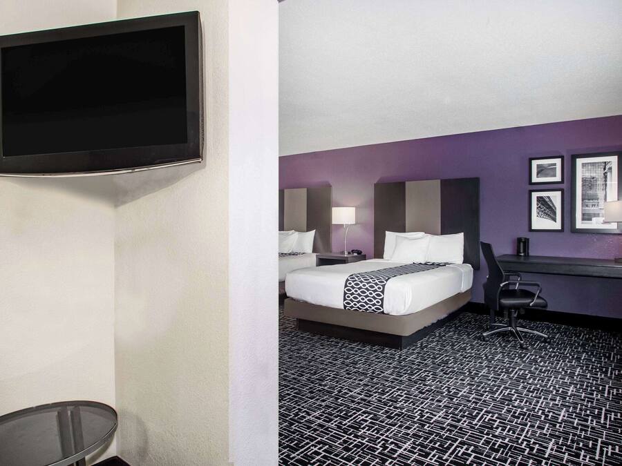 Quarto de hotel com cama, escrivaninha, cadeira e televisão na parede.