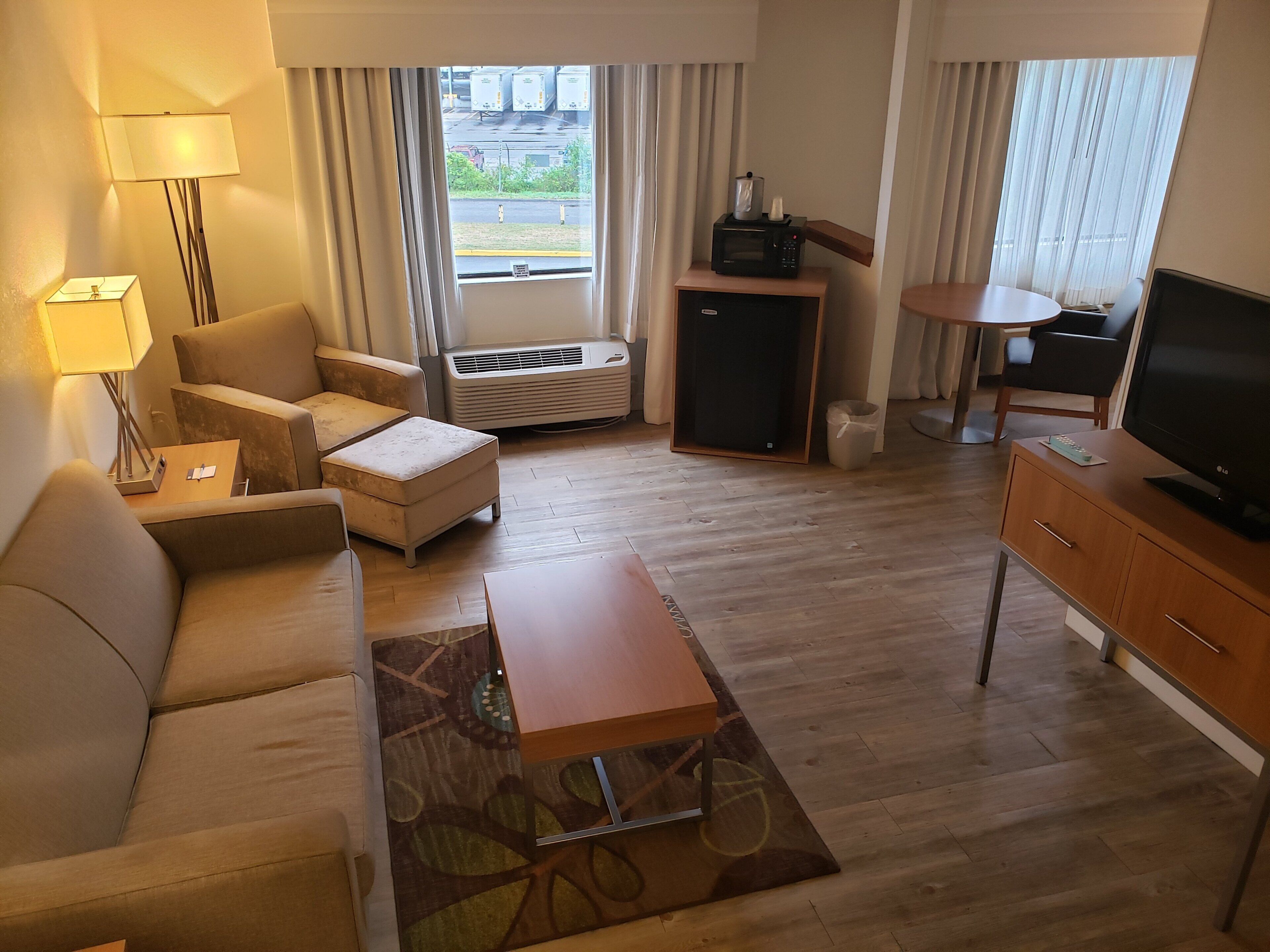 Quarto de hotel com sofá, poltrona, banqueta, mesa de centro, TV e uma mesinha de cabeceira com abajur.