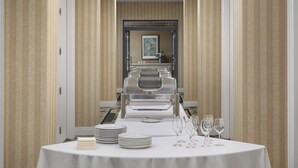 Uma área de refeições de hotel com serviço de buffet, uma mesa com pratos e copos empilhados, e um quadro na parede.