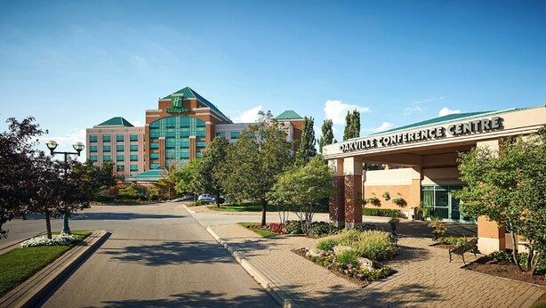 Um hotel Holiday Inn com fachada de tijolos e telhado verde, adjacente ao Centro de Convenções de Oakville.