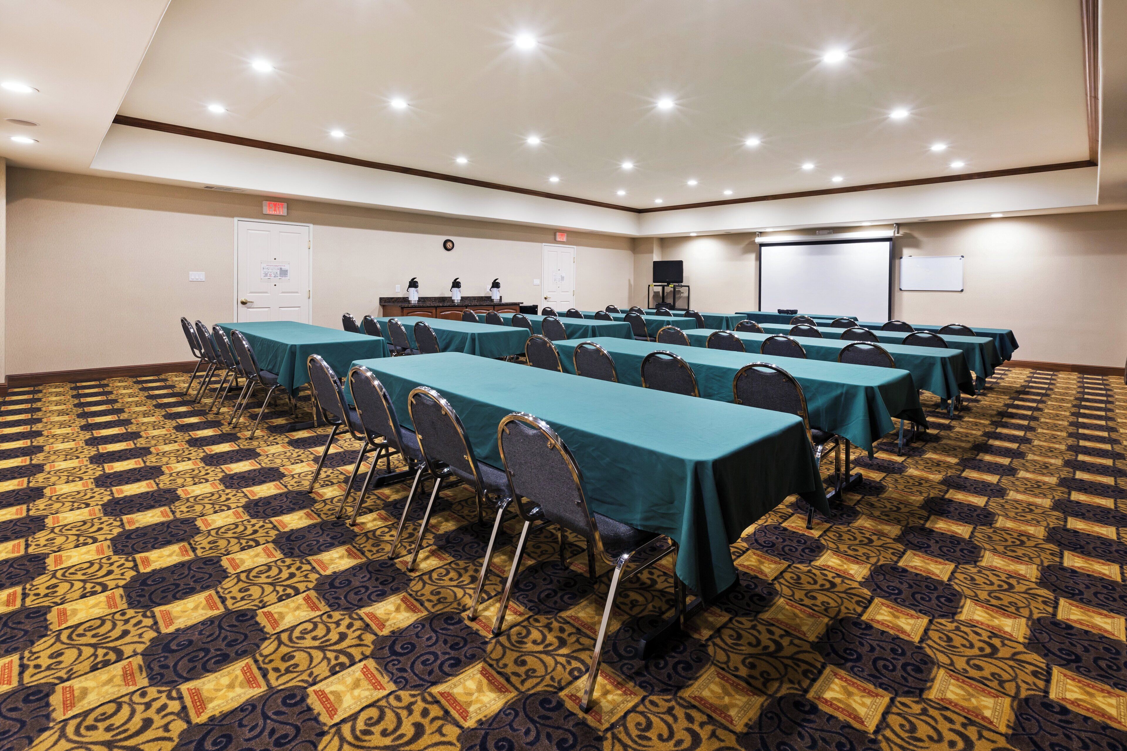 Sala de conferência com várias mesas e cadeiras, uma tela de projetor e piso coberto de carpete.