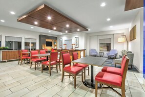 Uma área de refeições de hotel com cadeiras estofadas em vermelho, uma mesa central e um bar com televisão.