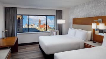 Quarto de hotel com duas camas, uma escrivaninha, uma lâmpada e vista para a paisagem urbana.