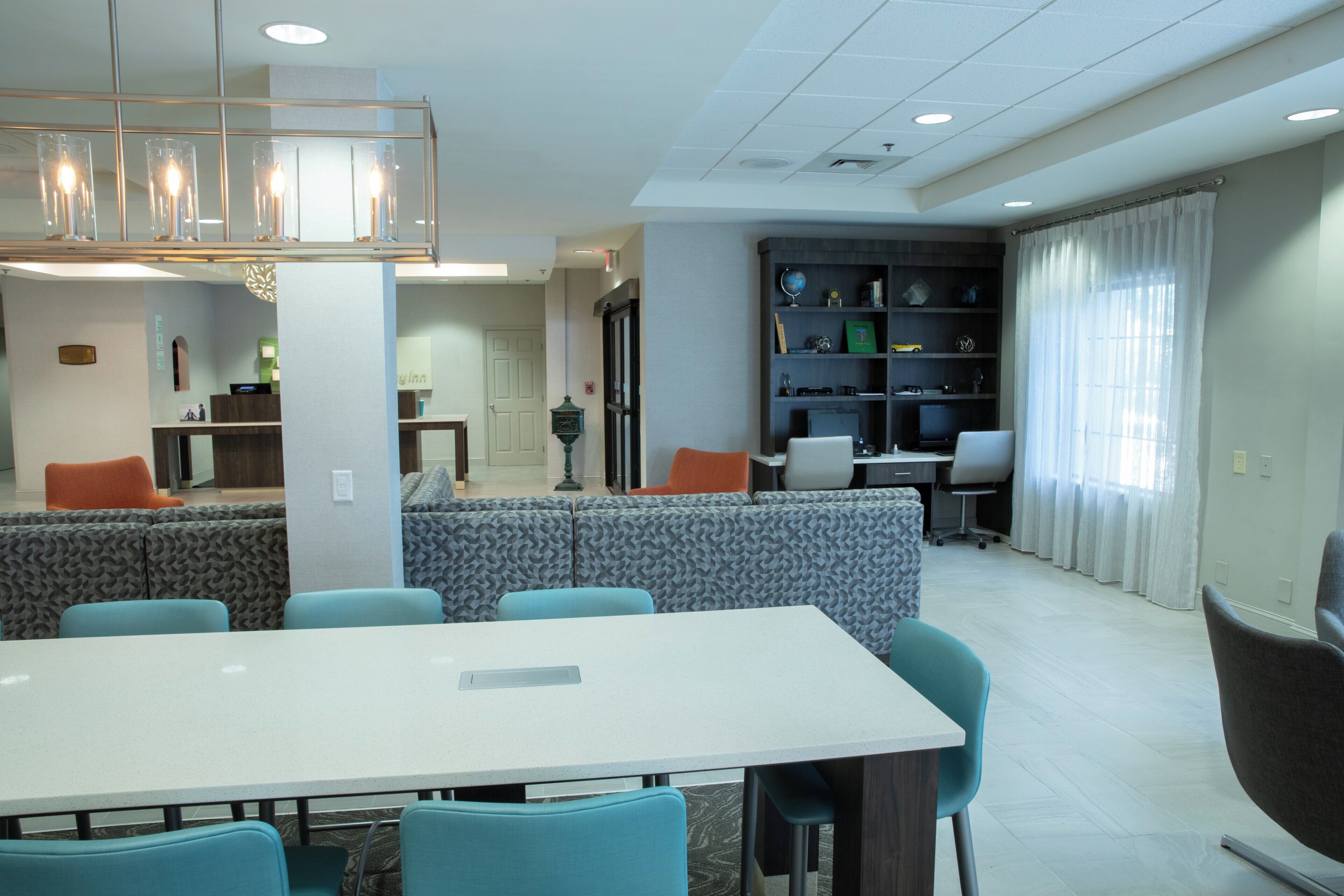 Uma área de refeições moderna com uma mesa comprida e cadeiras, um espaço de estar com almofadas estampadas e uma estante com diversos objetos.