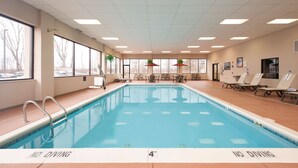 Piscina interna com placas de 'NÃO MERGULHAR', piso ladrilhado e janelas iluminadas pelo sol.