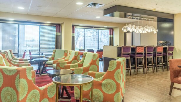 Um saguão de hotel com área de estar com cadeiras estampadas em laranja e verde, uma mesa de vidro e um bar com cadeiras roxas.