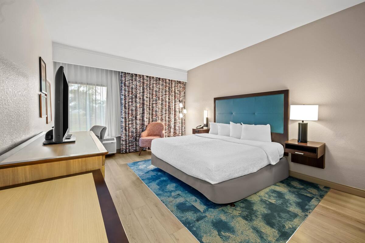 Quarto de hotel com cama, escrivaninha, cadeira e televisão.