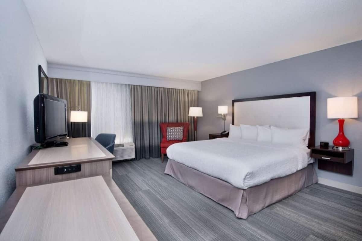 Quarto de hotel com cama, escrivaninha com televisão, cadeira, luminária vermelha e mesa de cabeceira com abajur.